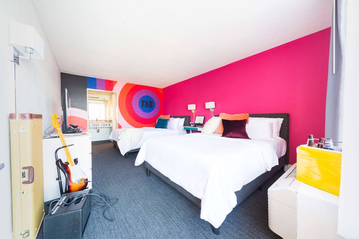 Quarto de hotel com duas camas, um violão, um minibar e um design de parede colorido.