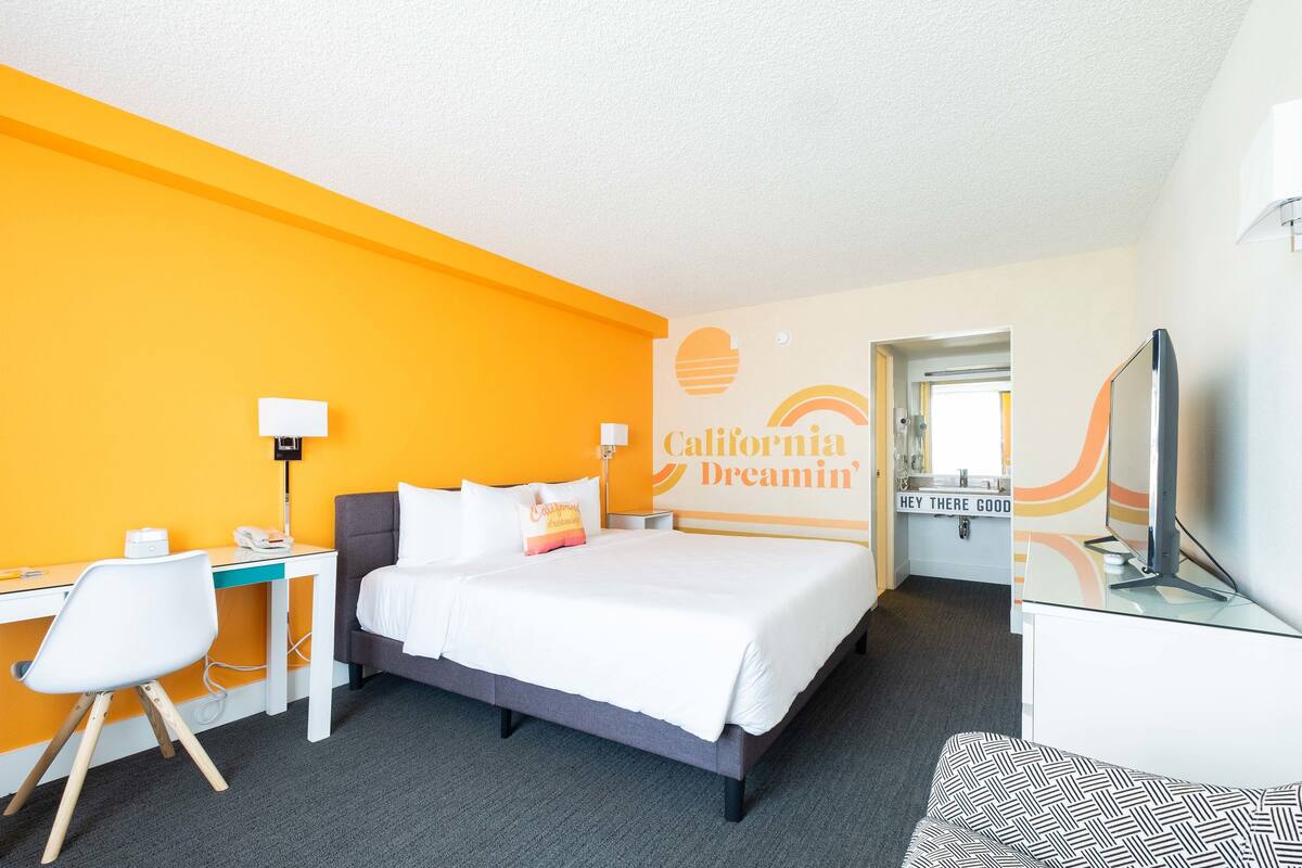 Quarto de hotel com cama, escrivaninha, cadeira, televisão e um quadro com a inscrição 'California Dreamin'.