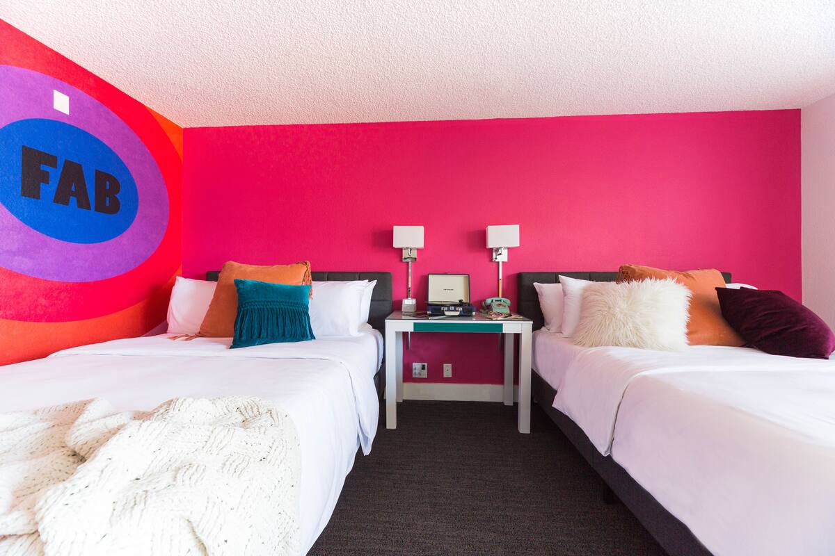 Quarto de hotel com duas camas, uma mesa de cabeceira com luminárias e uma parede com o letreiro 'FAB'.