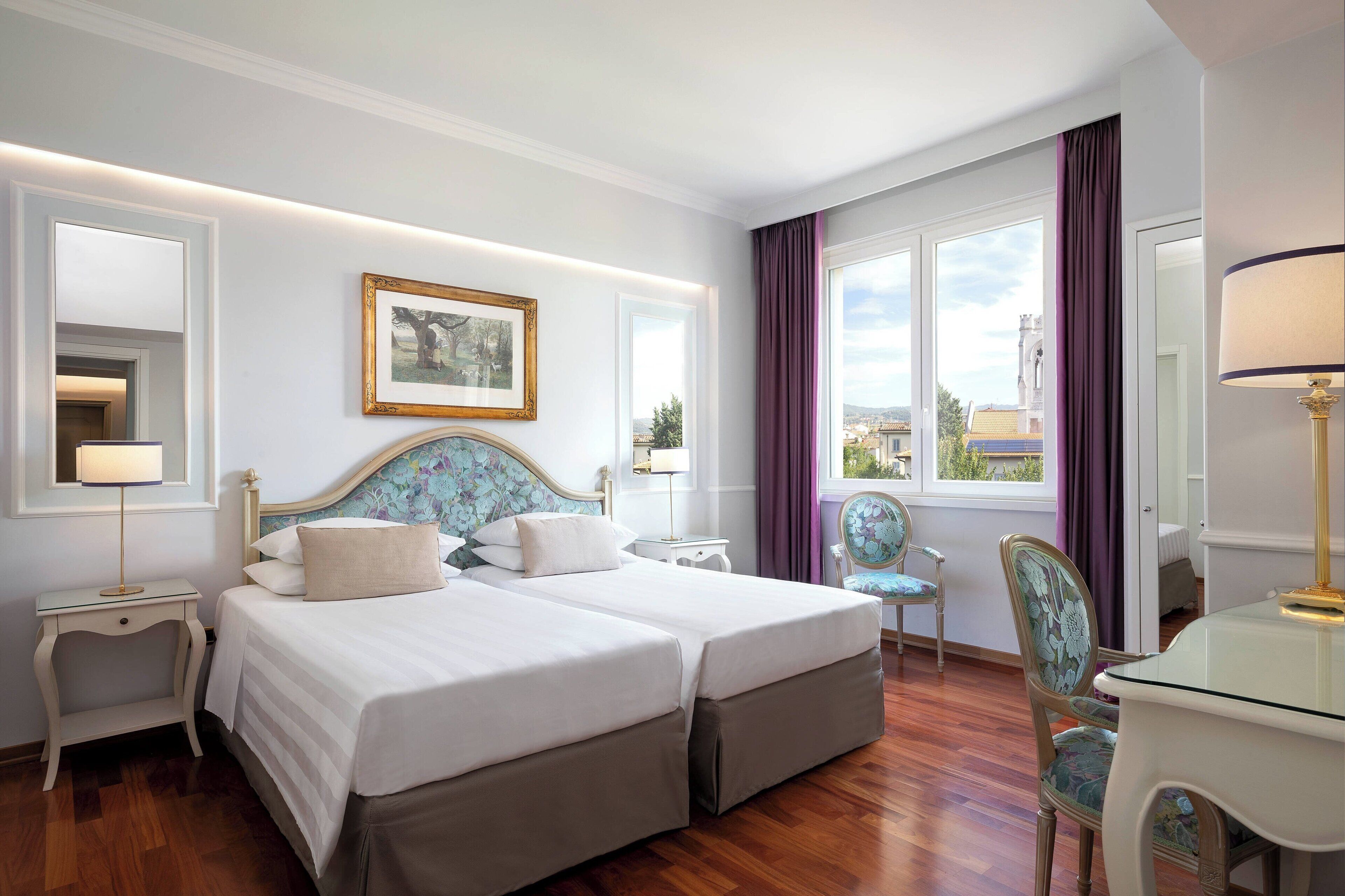Quarto de hotel com duas camas, uma mesa, uma cadeira, janela com vista e um quadro na parede.