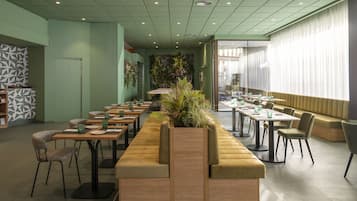 Interior de restaurante moderno com mesas comunitárias alongadas, assentos acolchoados e uma parede verde decorada com plantas.
