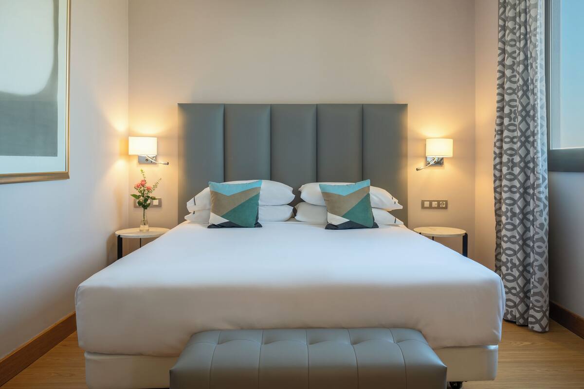 Quarto de hotel bem organizado, com uma cama grande, duas luminárias de cabeceira, uma mesa de cabeceira com um vaso e uma janela com cortinas estampadas.