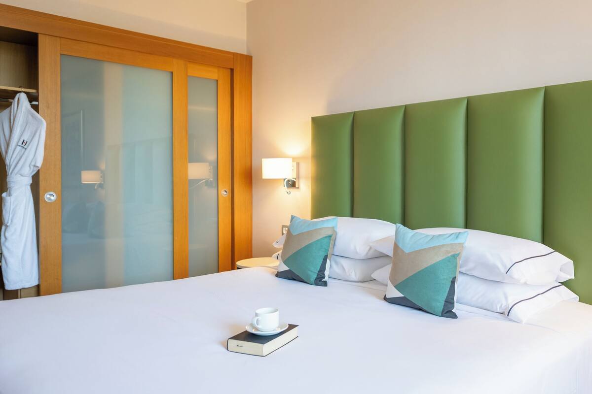 Quarto de hotel com uma cama, um roupão pendurado na porta e uma chávena sobre um livro.