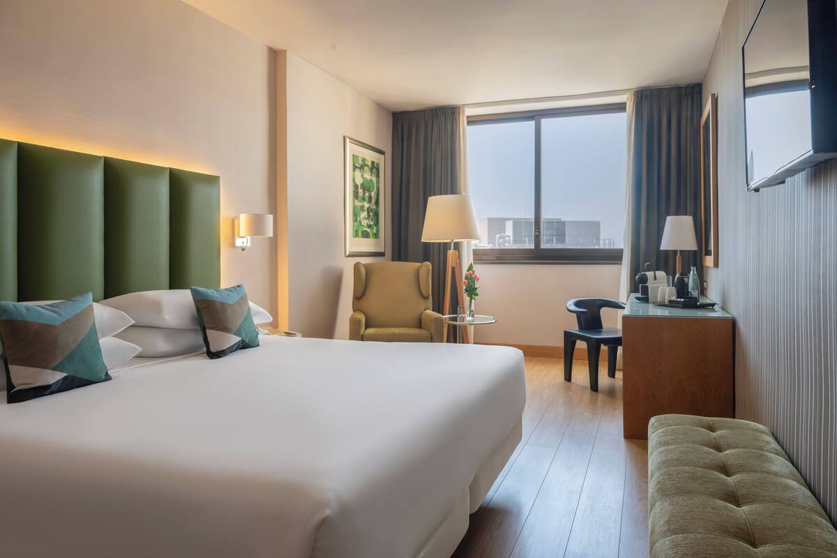 Quarto de hotel com uma cama grande, uma cadeira, uma mesinha, uma escrivaninha e uma janela com cortinas.