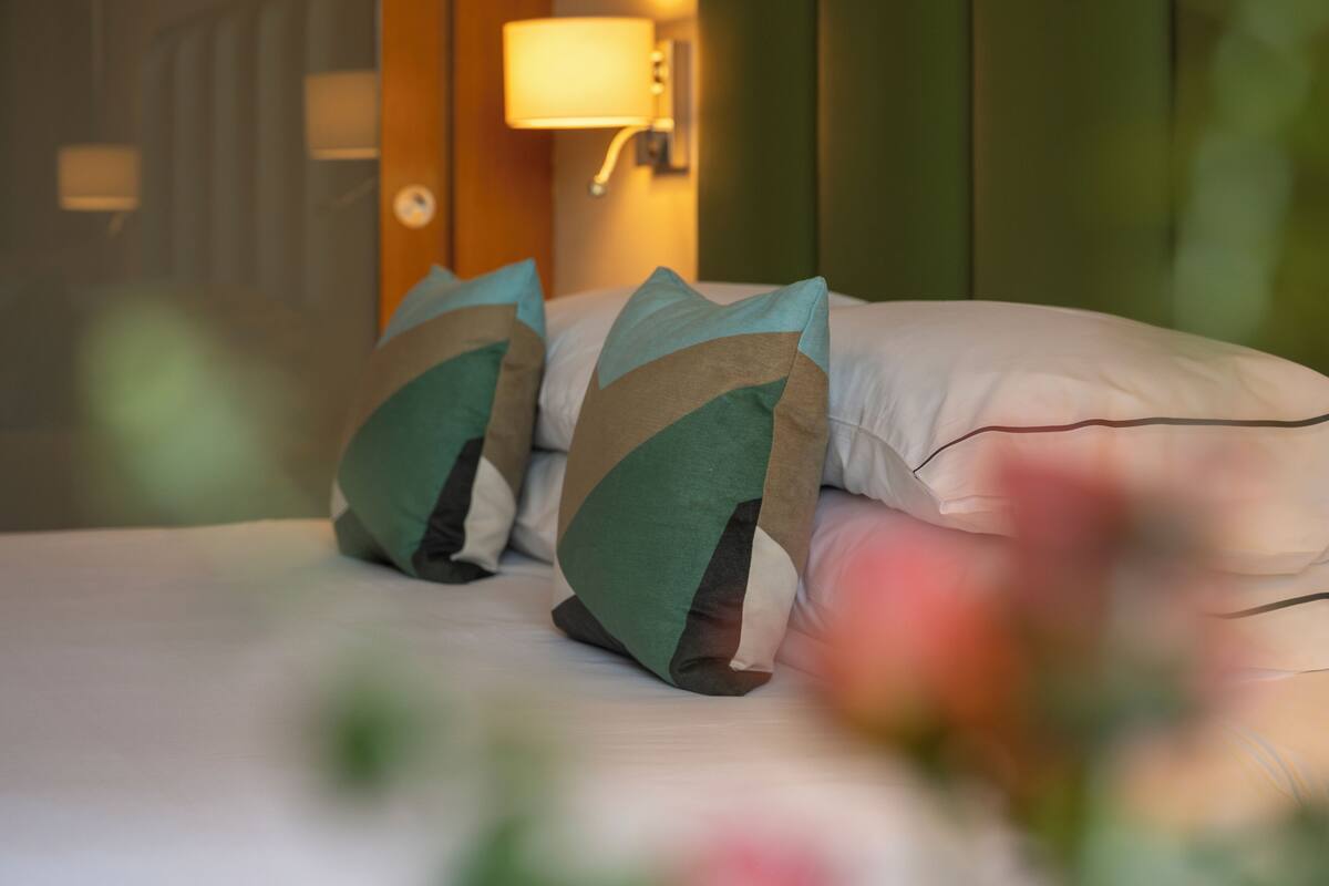 Quarto de hotel com cama, almofadas e uma luminária fixada na parede.