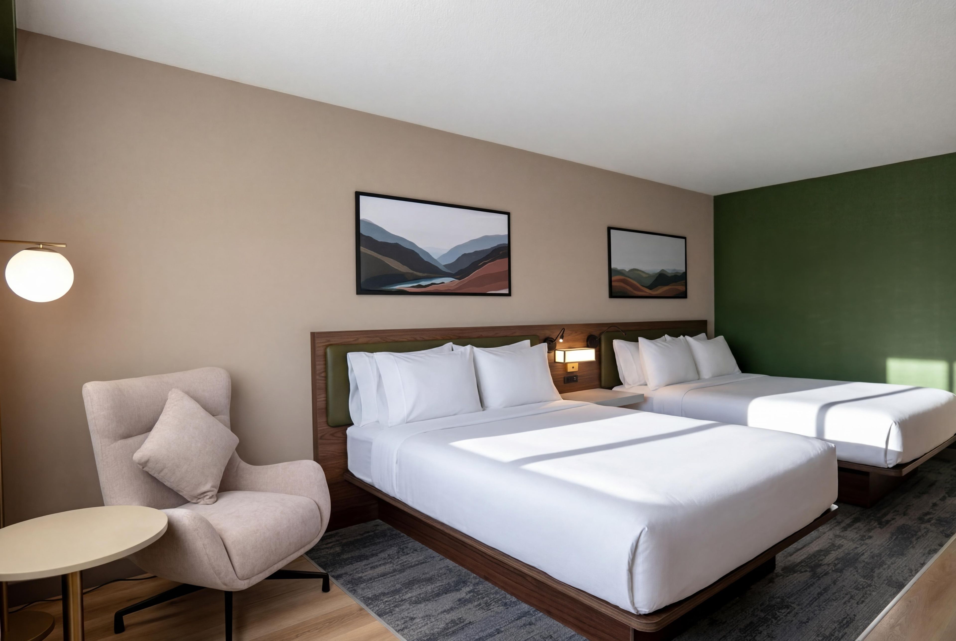 Quarto de hotel com duas camas, uma cadeira, uma mesinha e duas luminárias fixadas na parede.