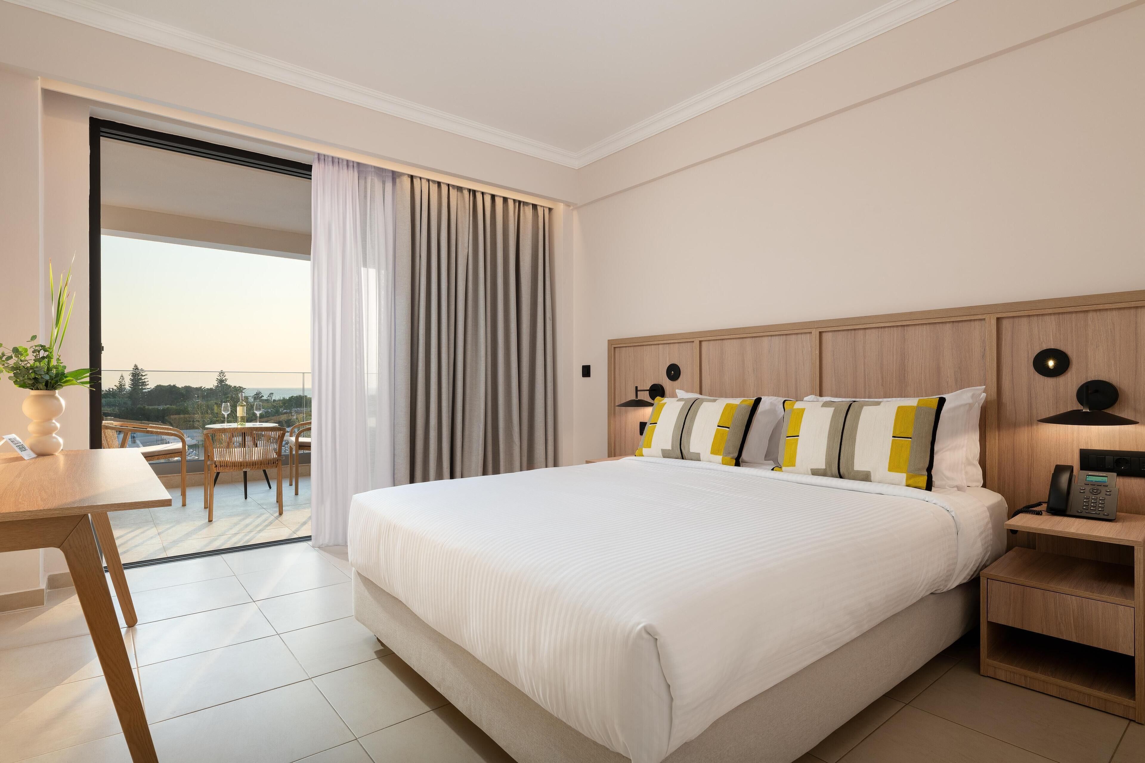 Quarto de hotel com uma cama grande, uma mesinha de cabeceira de madeira, uma escrivaninha com cadeira e uma varanda com mesa e cadeiras.