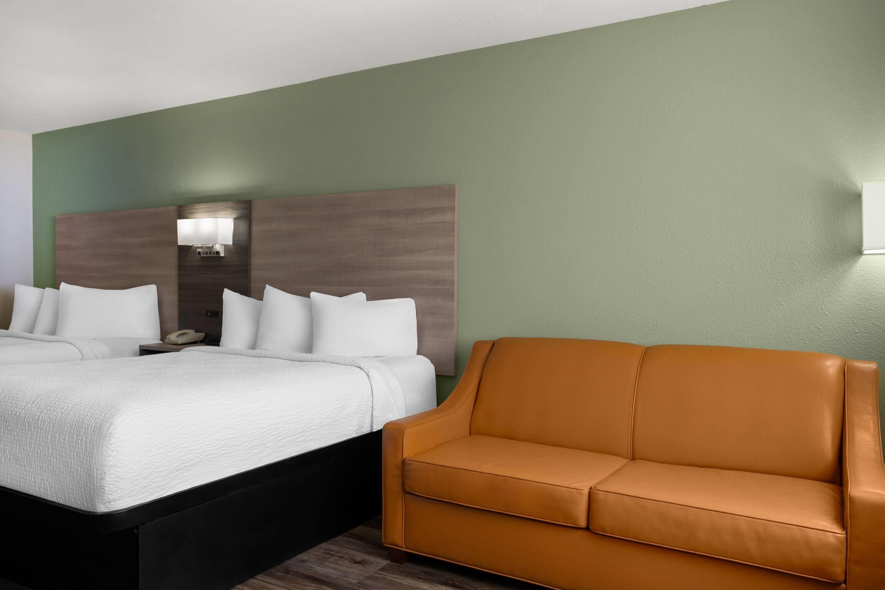 Quarto de hotel com cama, sofá, mesa de cabeceira com abajur e luminária na parede.