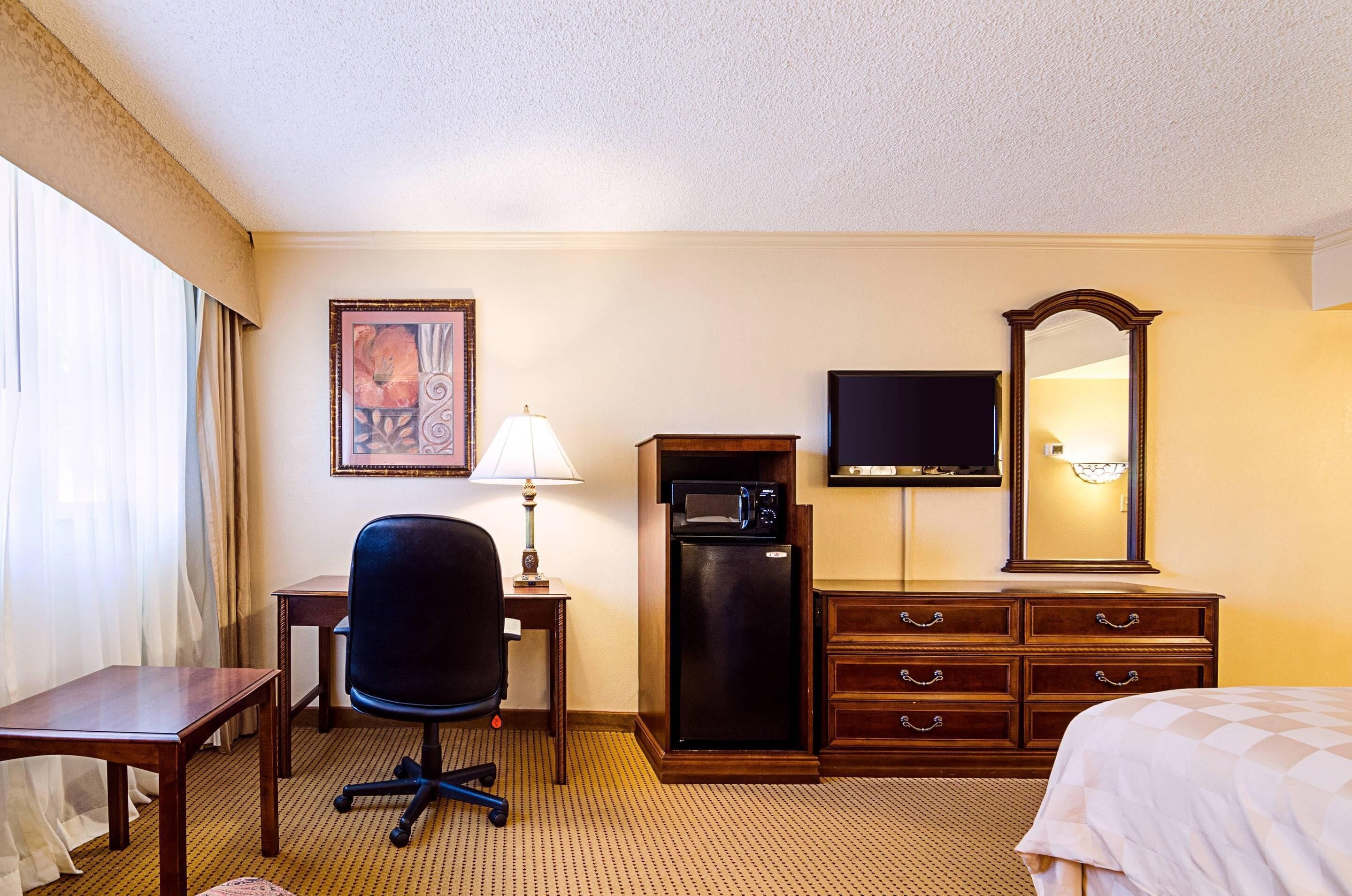 Quarto de hotel com cama, escrivaninha, cadeira, televisão, micro-ondas, penteadeira e espelho.