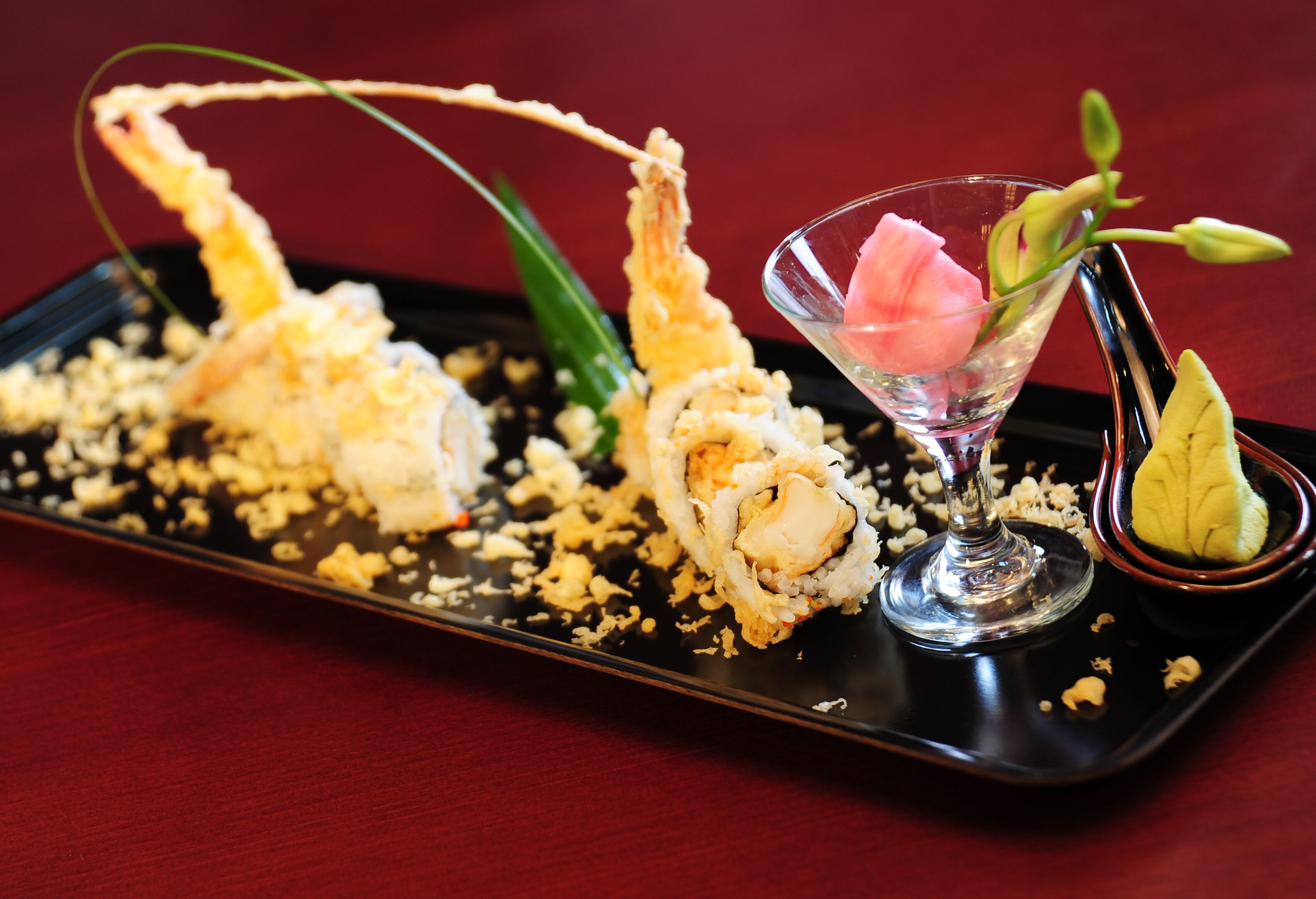 Um prato de sushi com um copo de bebida rosa e uma flor decorativa sobre um tabuleiro preto.