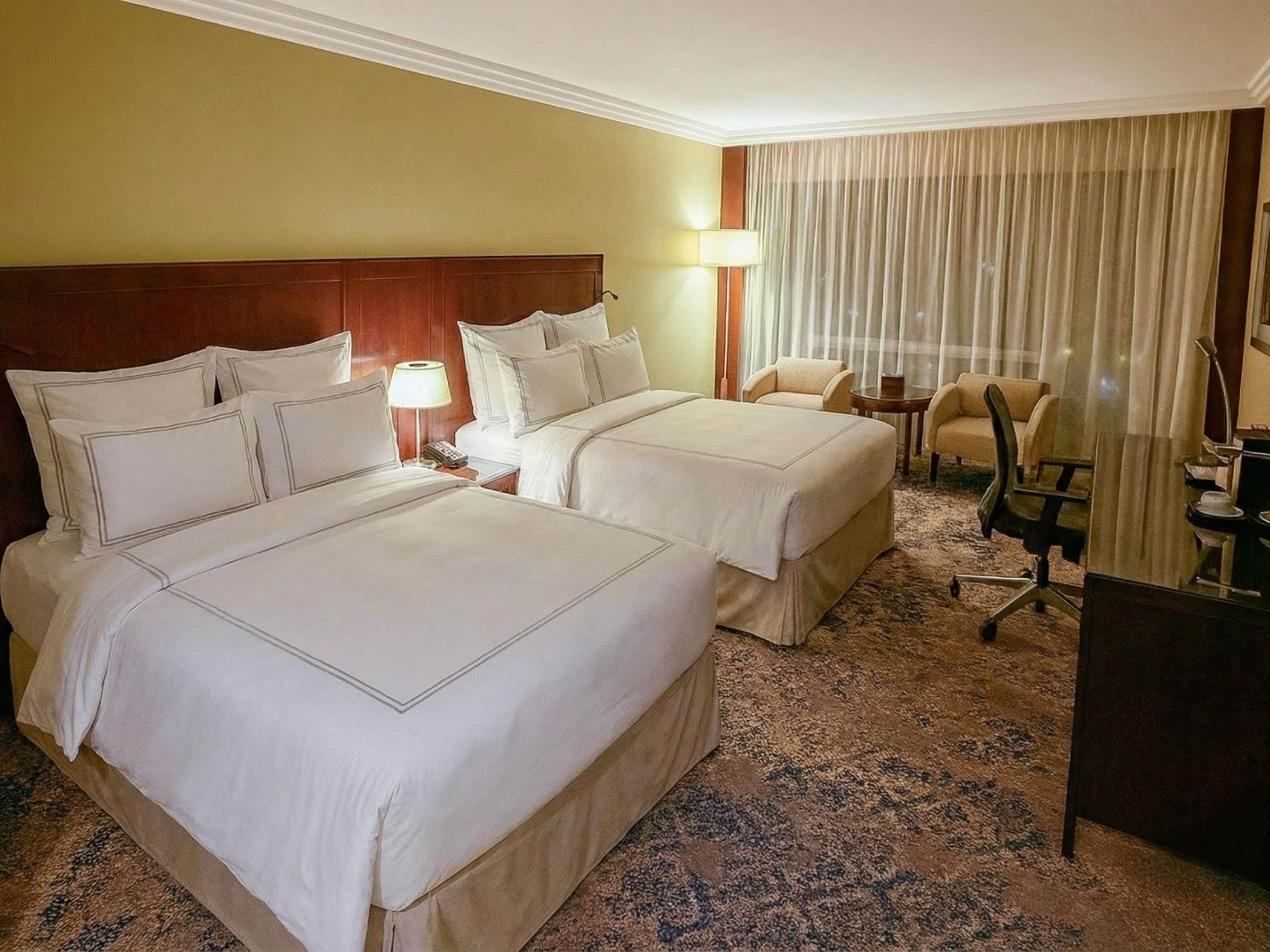 Quarto de hotel com duas camas, uma escrivaninha, uma cadeira e uma janela grande com cortinas.