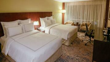Quarto de hotel com duas camas, uma escrivaninha, uma cadeira e uma janela grande com cortinas.