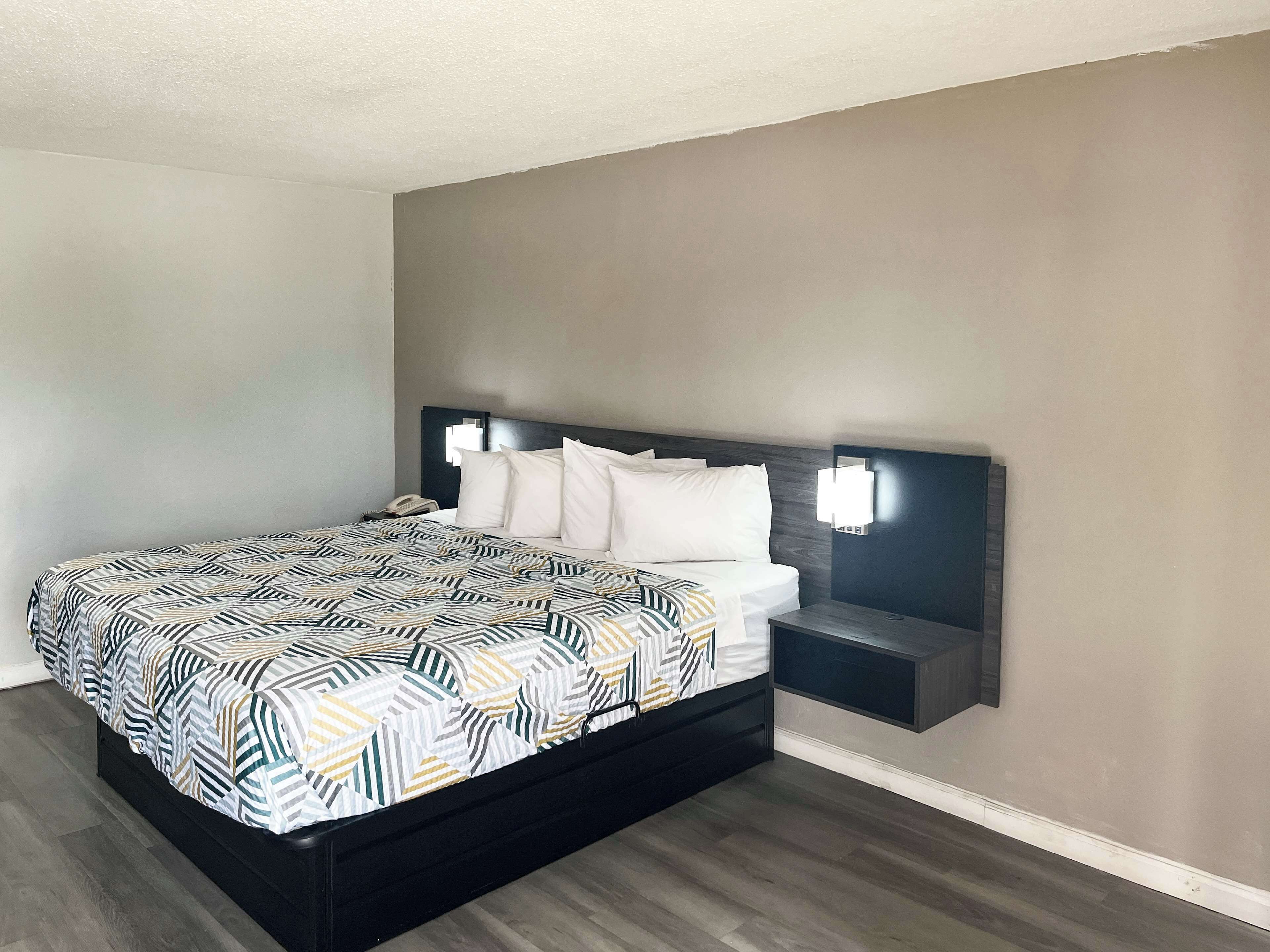 Quarto de hotel com cama, mesa de cabeceira e luminária fixada na parede.