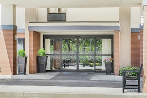 Entrada de hotel com porta de vidro, plantas em vasos e um banco.