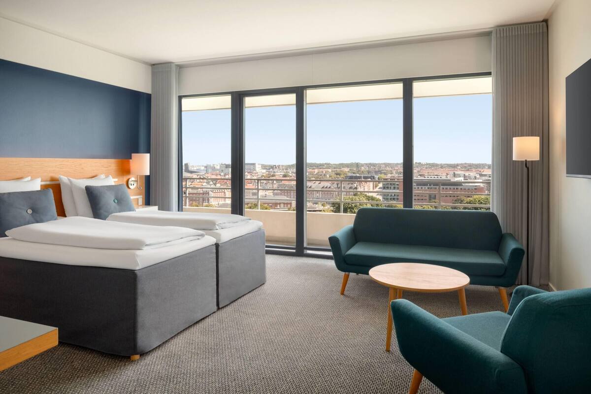 Quarto de hotel com uma cama grande, um sofá, uma mesa pequena e vista para a cidade.