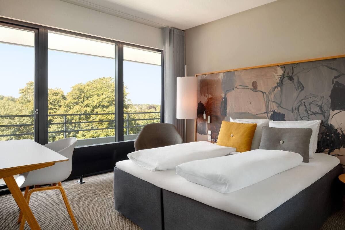 Quarto de hotel com uma grande janela, cama com roupa de cama branca, escrivaninha e uma cadeira.