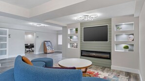 Uma sala de estar moderna com uma televisão de tela plana grande, lareira e uma mesa de centro redonda. Há sofás azuis e um tapete pequeno no chão.