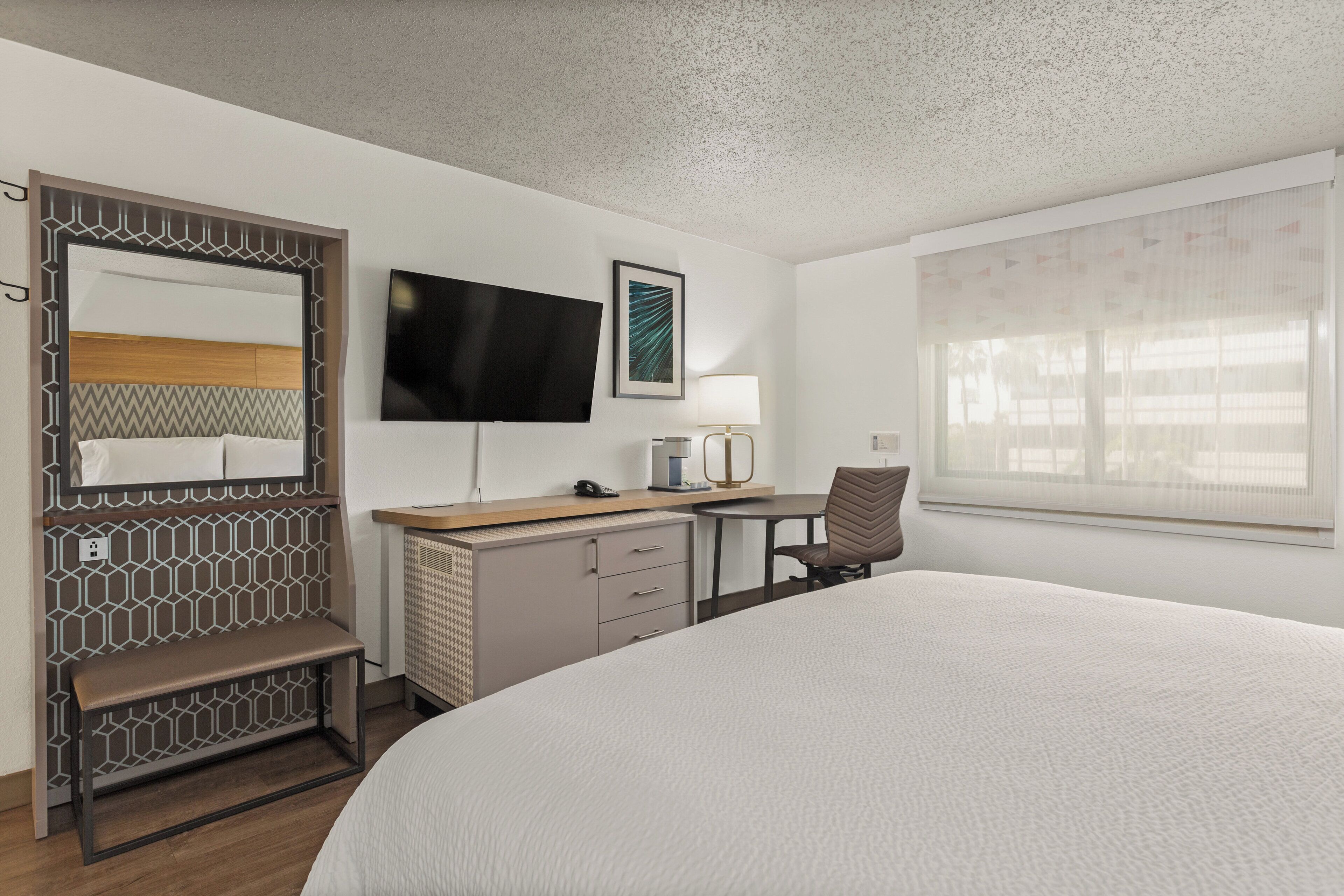 Quarto de hotel com uma cama grande, uma escrivaninha, uma cadeira, uma televisão e uma janela com cortinas.