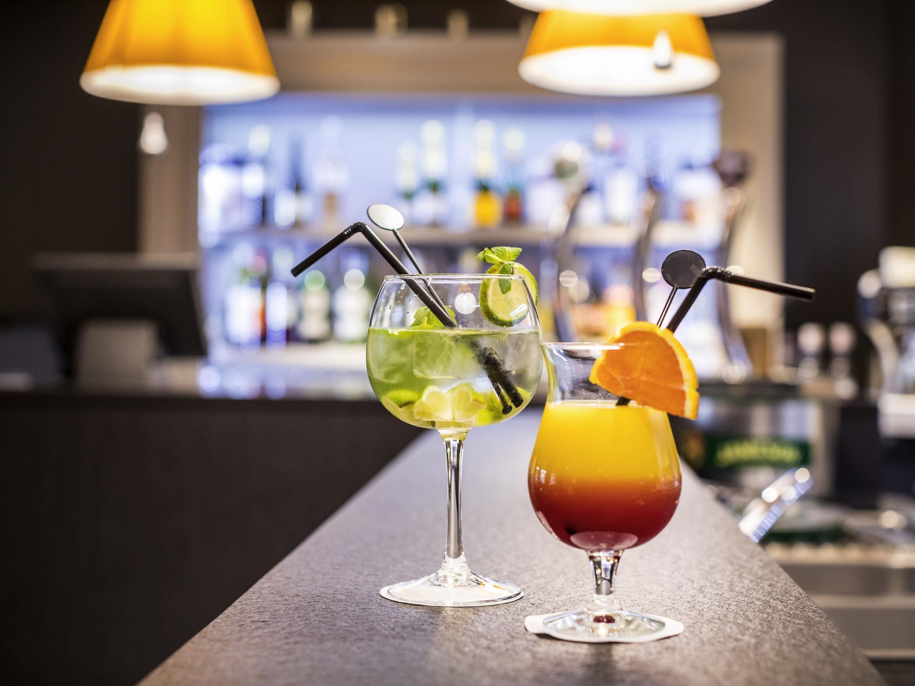 Dois cocktails sobre um balcão com fundo desfocado de garrafas e área de bar.