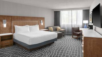Quarto de hotel com uma cama grande, uma escrivaninha, uma cadeira, uma televisão e uma janela com cortinas.