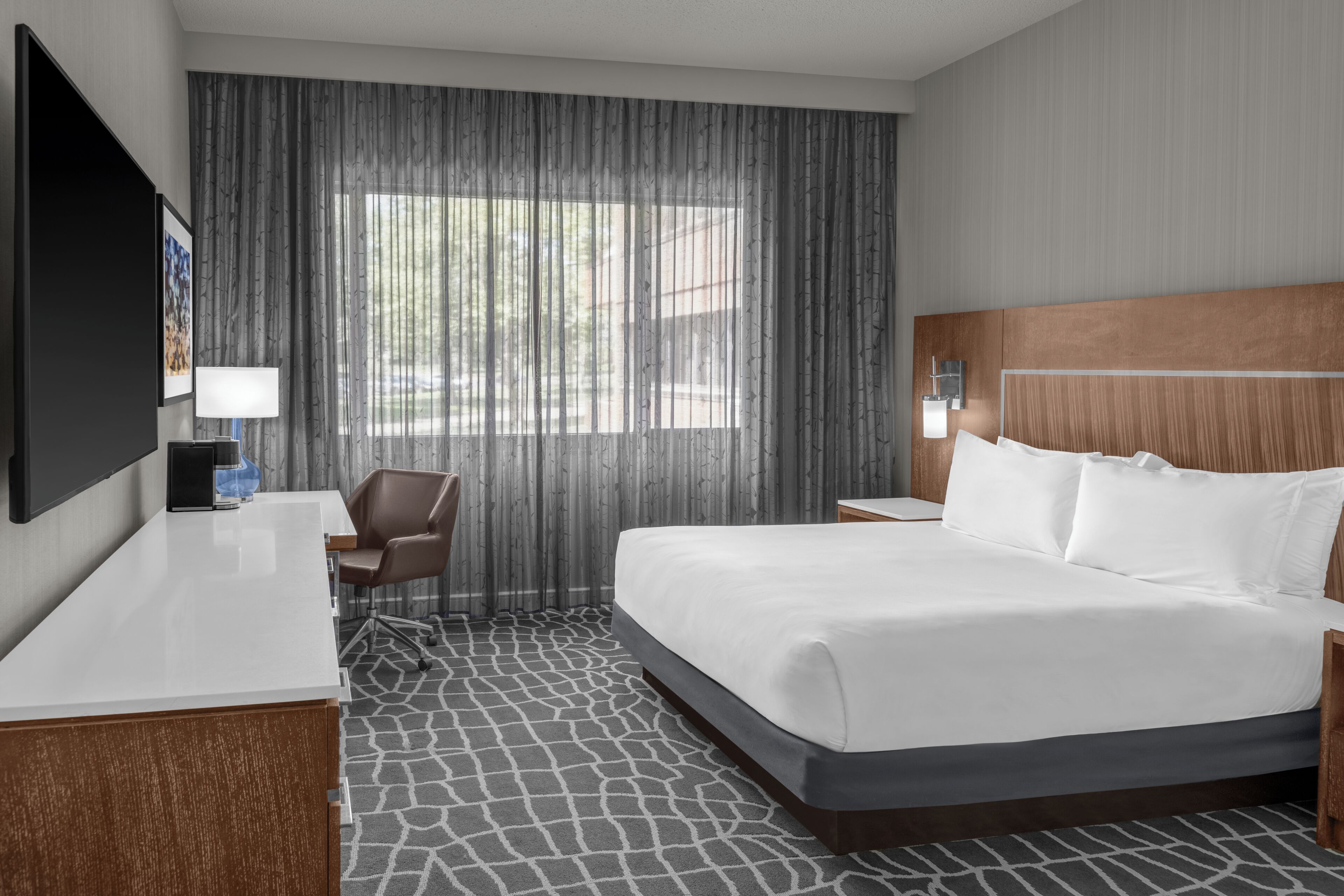 Quarto de hotel com cama, escrivaninha, cadeira, televisão e janela com cortinas.