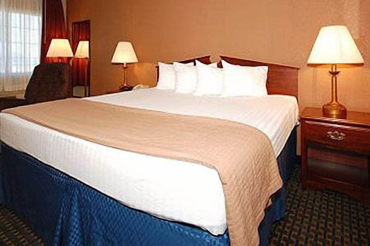Quarto de hotel com uma cama grande, duas luminárias de cabeceira, um painel de cabeceira de madeira e uma janela com cortinas.