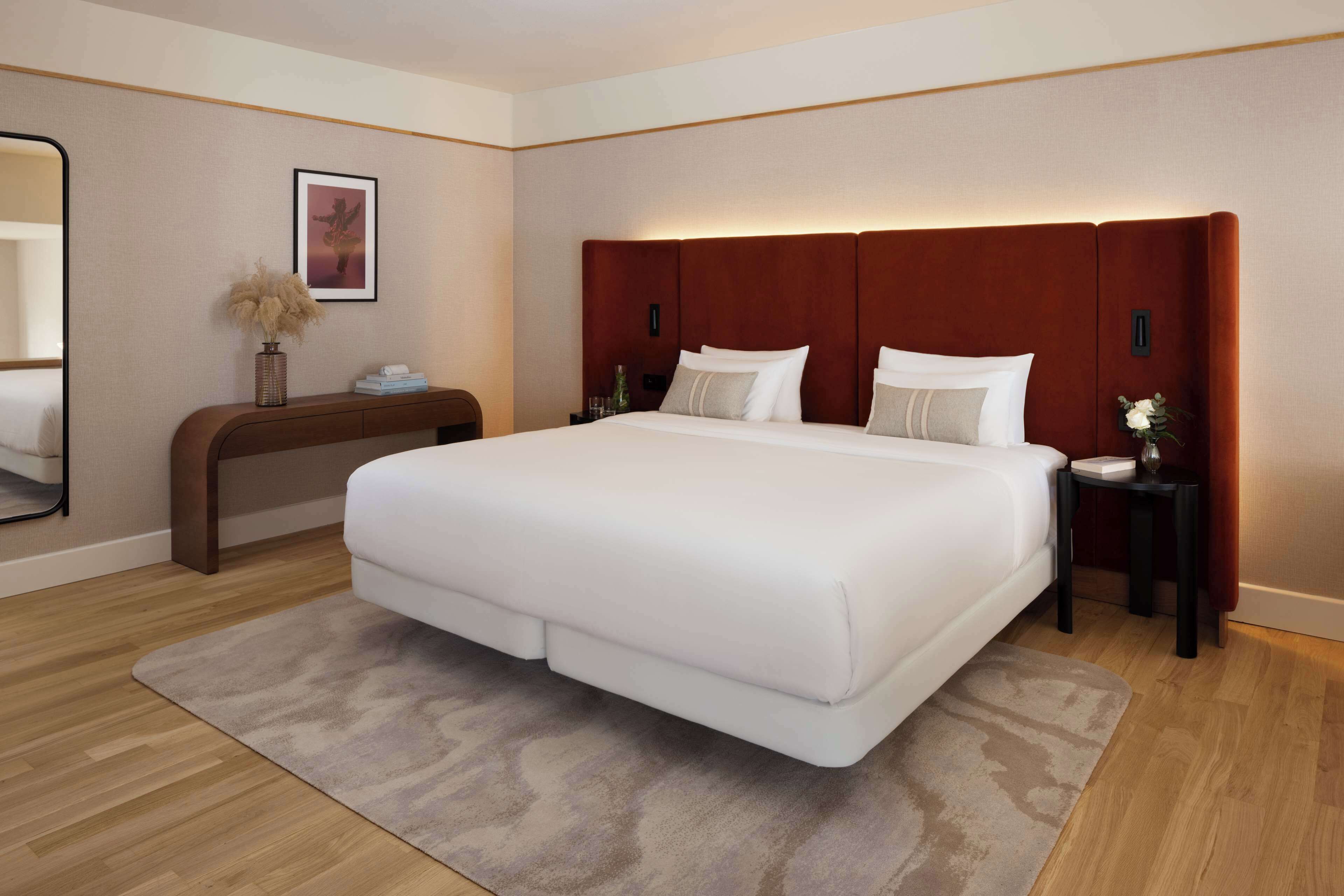 Quarto de hotel moderno com uma cama grande, uma mesa de cabeceira, um espelho, um banco e um quadro na parede.