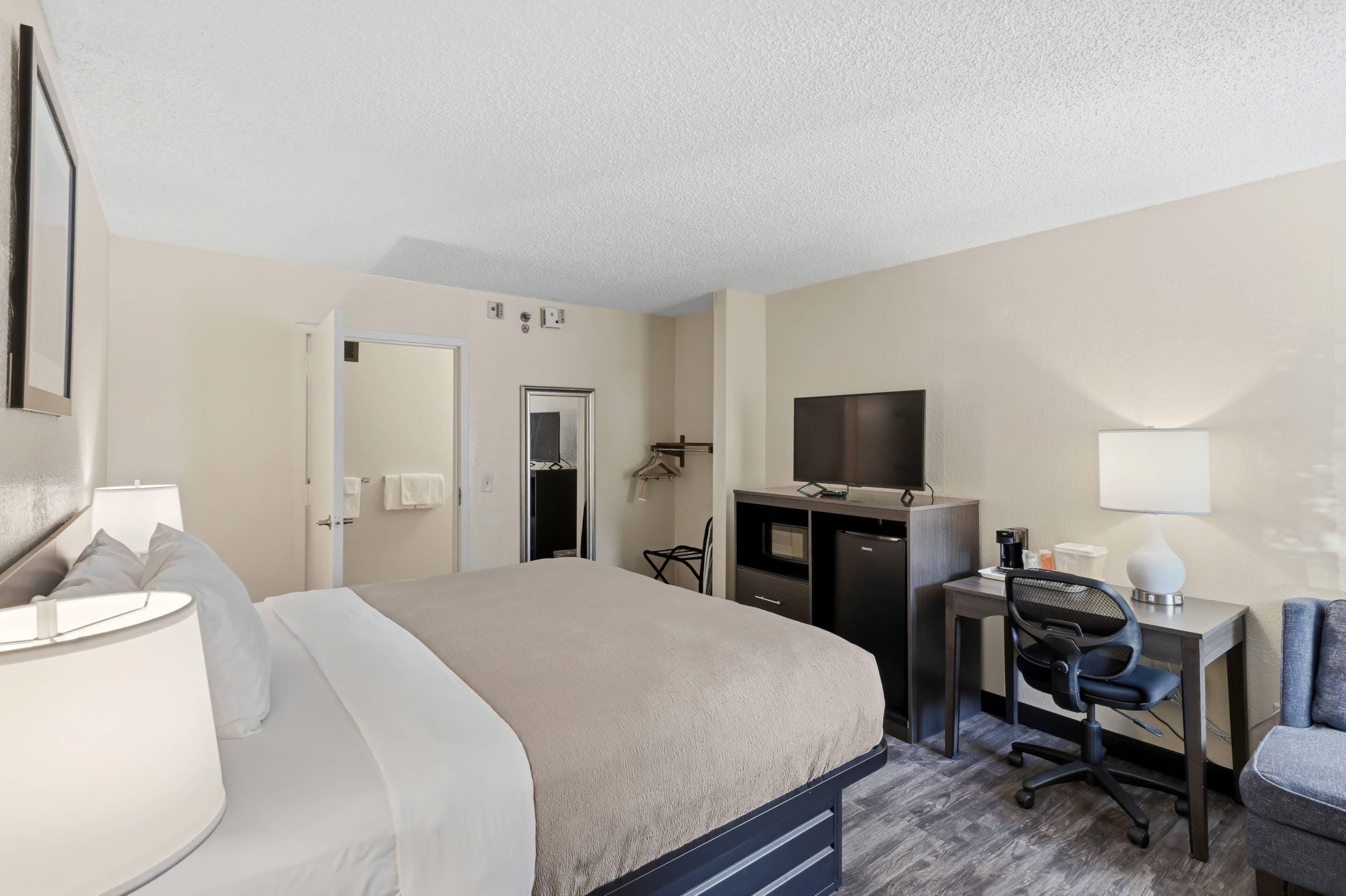 Quarto de hotel com cama, escrivaninha, cadeira, televisão e espelho.
