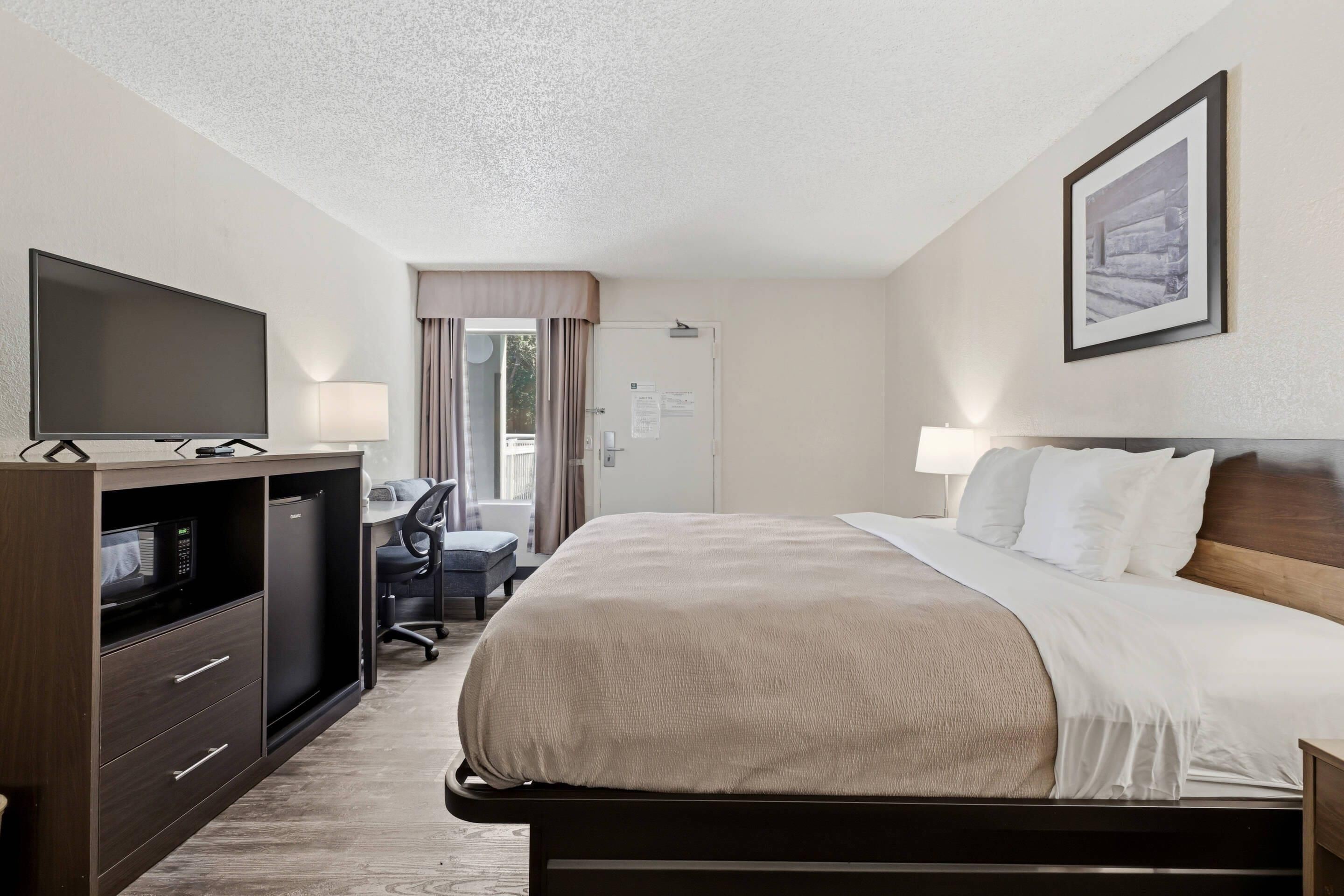 Quarto de hotel com cama, escrivaninha, cadeira, televisão e porta.