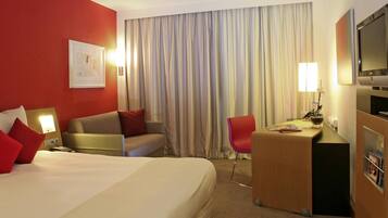 Quarto de hotel com cama, escrivaninha, cadeira, televisão e janela com cortinas.