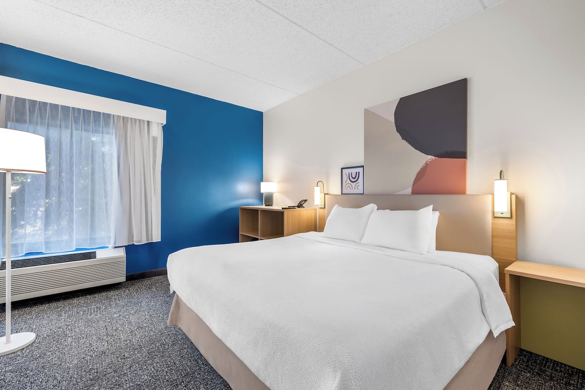 Quarto de hotel com uma cama grande, mesas de cabeceira, um abajur, uma janela com cortinas e uma obra de arte pendurada na parede.