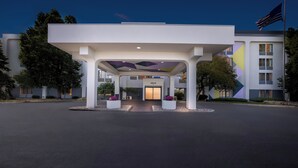 Entrada moderna de hotel com área coberta, uma bandeira e um edifício com fachada geométrica.
