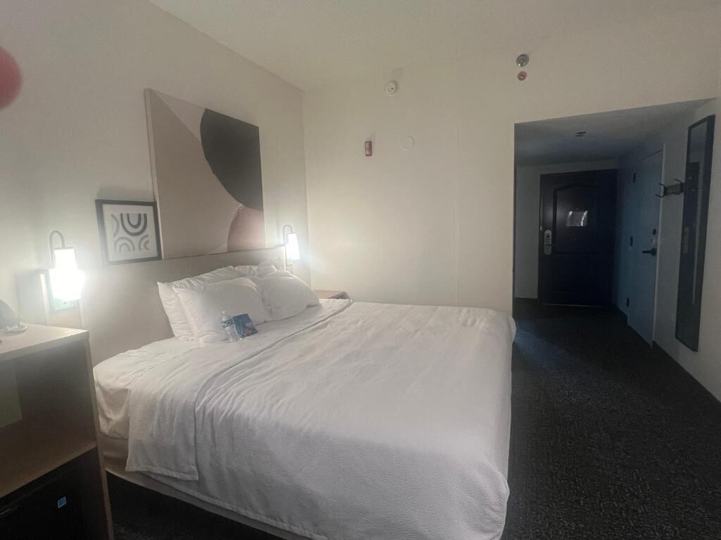 Quarto de hotel com uma cama, mesas de cabeceira, uma luminária na parede e uma porta que dá para outro cômodo.