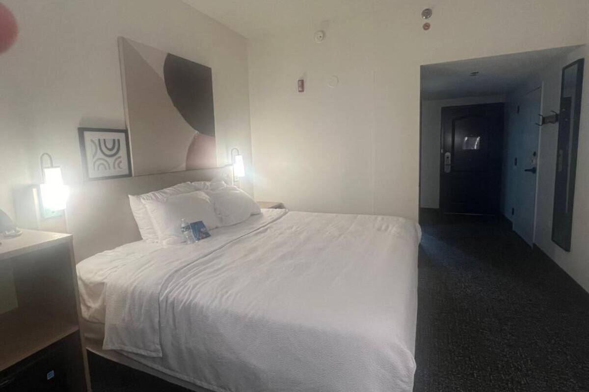 Quarto de hotel com uma cama grande, mesas de cabeceira, uma obra de arte na parede e uma porta que dá para outro quarto.