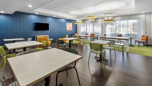 Uma sala de jantar moderna com várias mesas e cadeiras, uma TV de tela plana na parede e grandes janelas com cortinas leves.