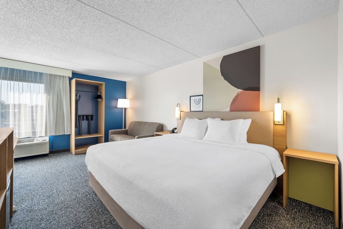 Quarto de hotel com uma cama grande, uma escrivaninha, uma cadeira, um guarda-roupa e uma janela com cortinas.