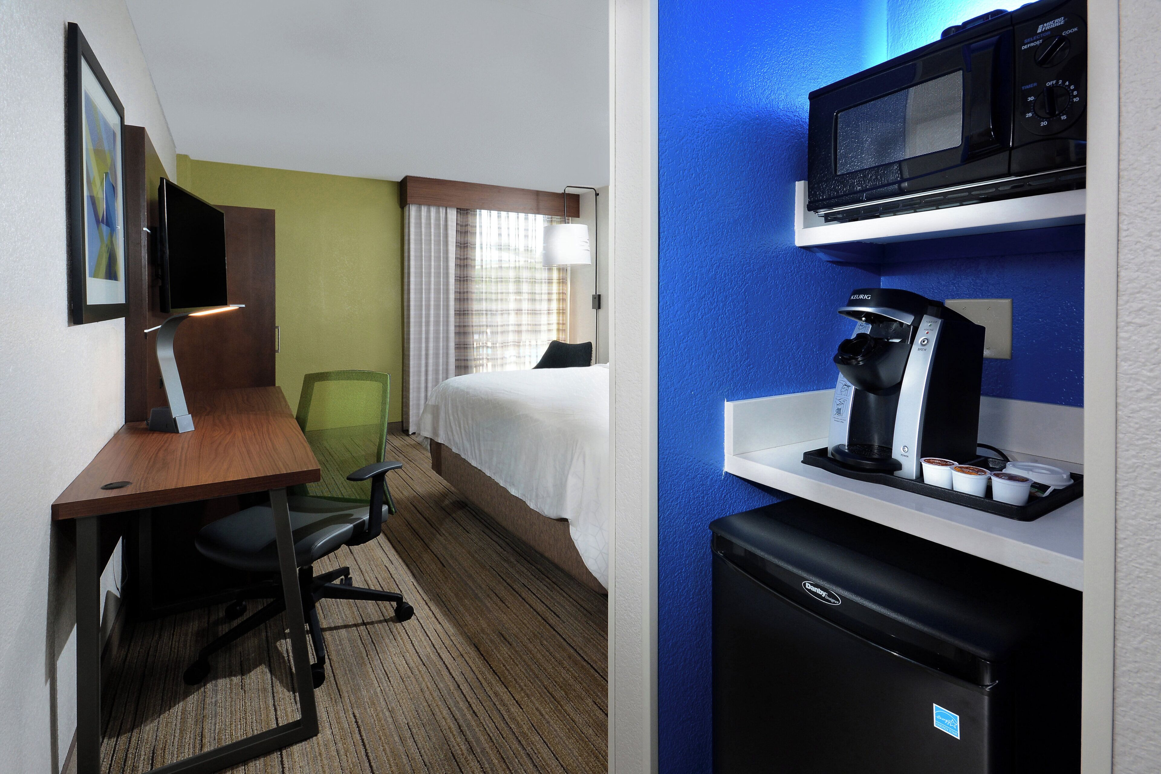Quarto de hotel com uma cama, uma escrivaninha com cadeira, cozinha compacta com cafeteira e uma televisão na parede.