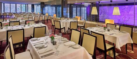 Salão de banquetes com várias mesas preparadas para refeições, grandes janelas e design de interiores moderno.