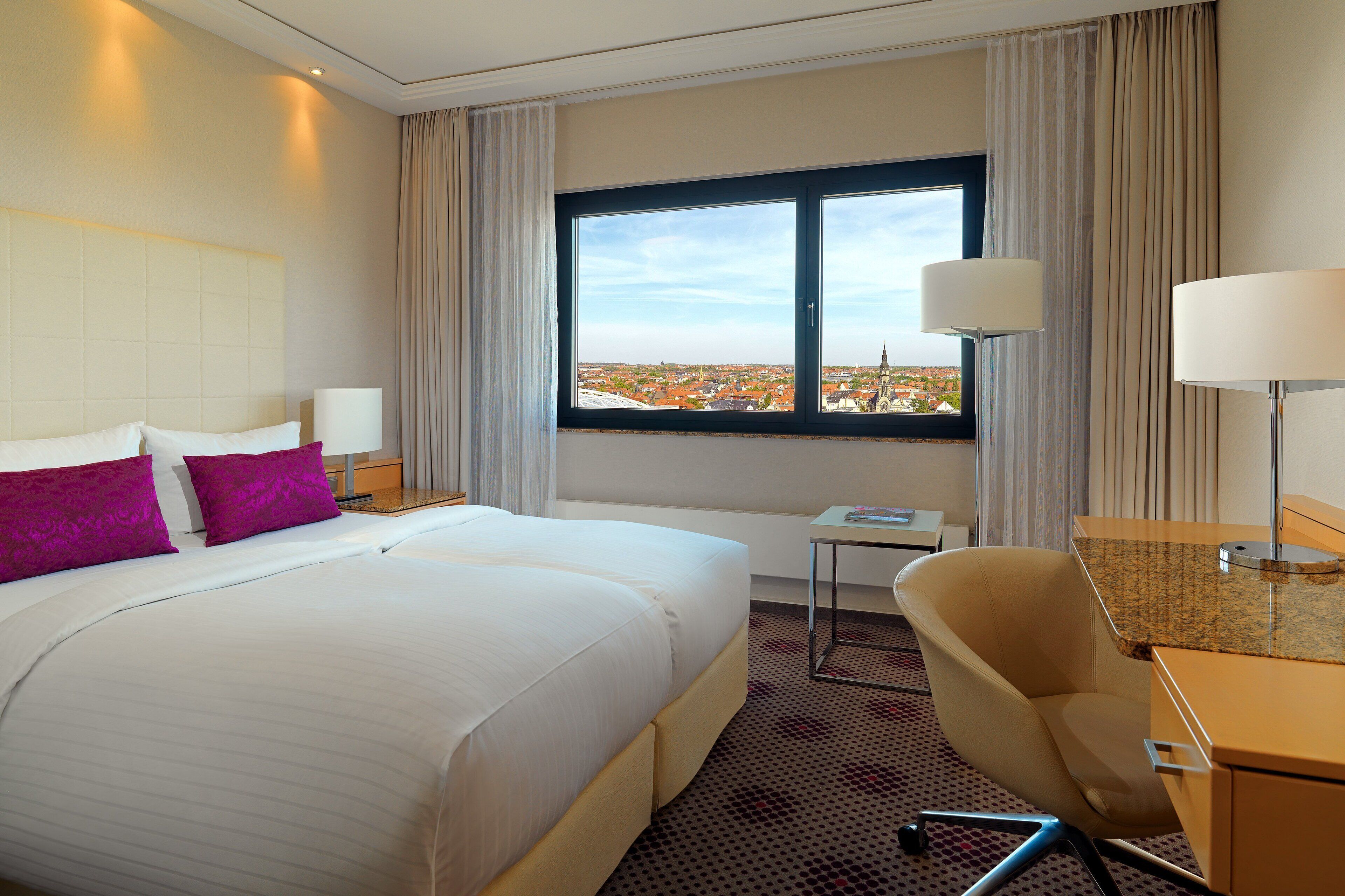 Quarto de hotel com uma cama grande, uma escrivaninha, uma cadeira e uma janela com vista para a cidade.