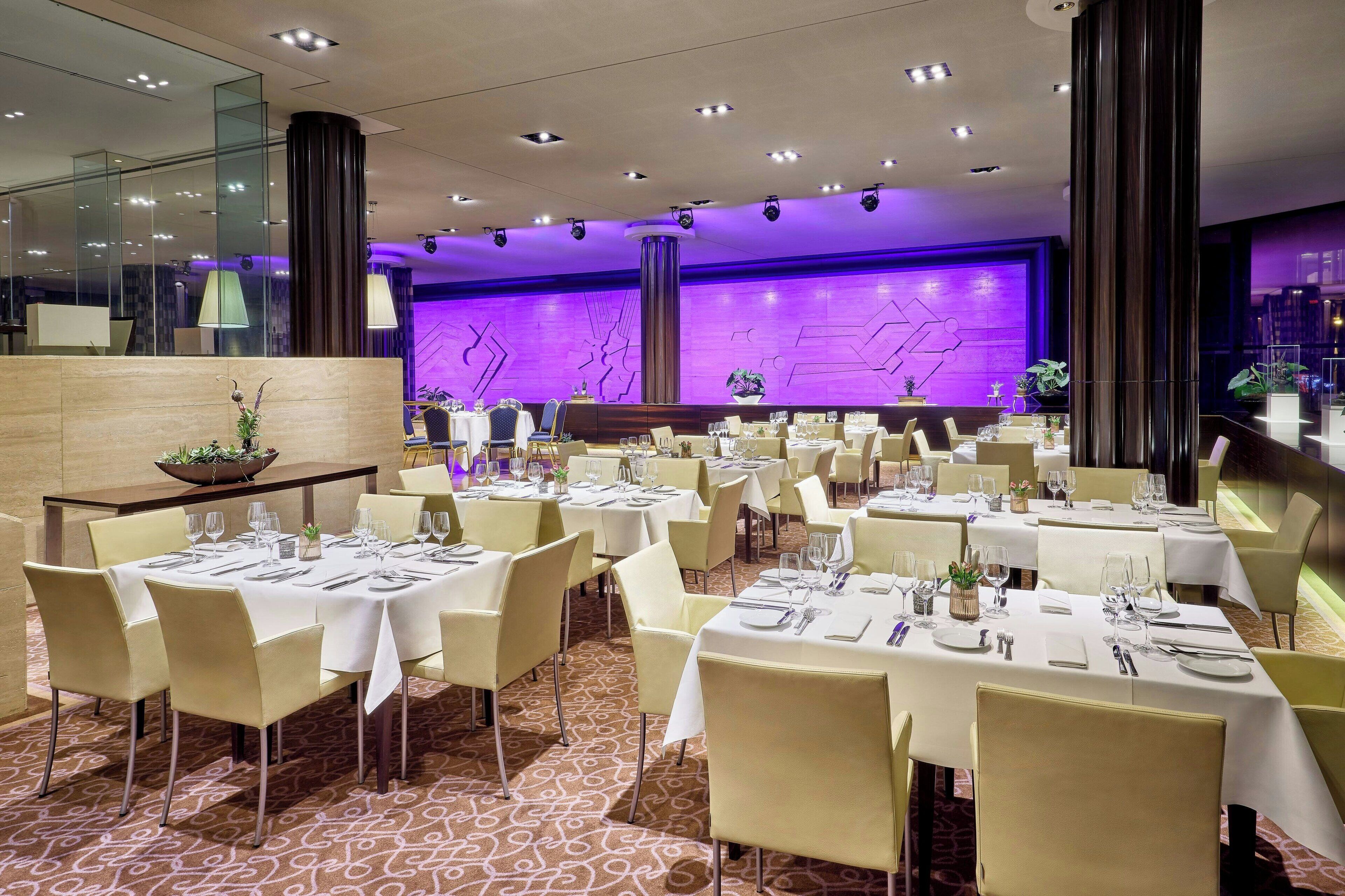 Uma área de refeições com várias mesas preparadas para hóspedes, apresentando um interior moderno com uma parede iluminada em roxo e um grande pilar.