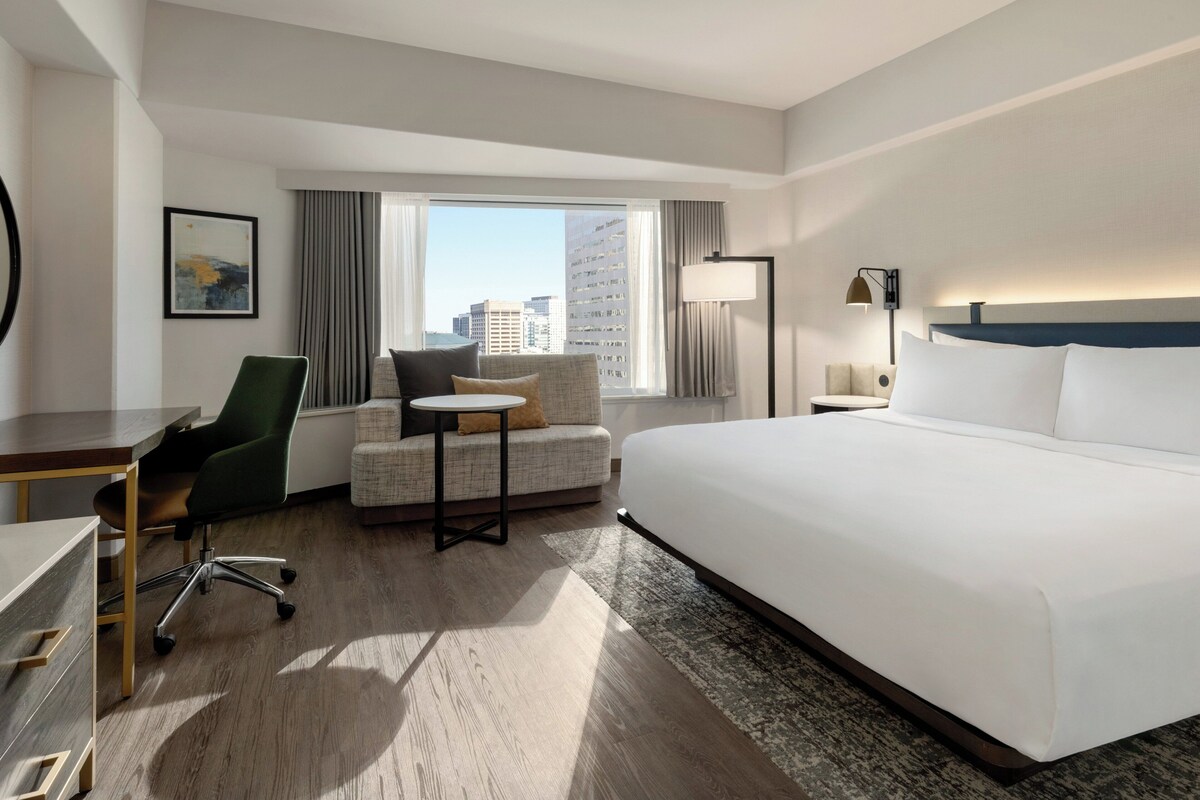 Quarto de hotel com uma cama grande, uma escrivaninha, uma cadeira, uma mesinha e vista para a cidade pela janela.
