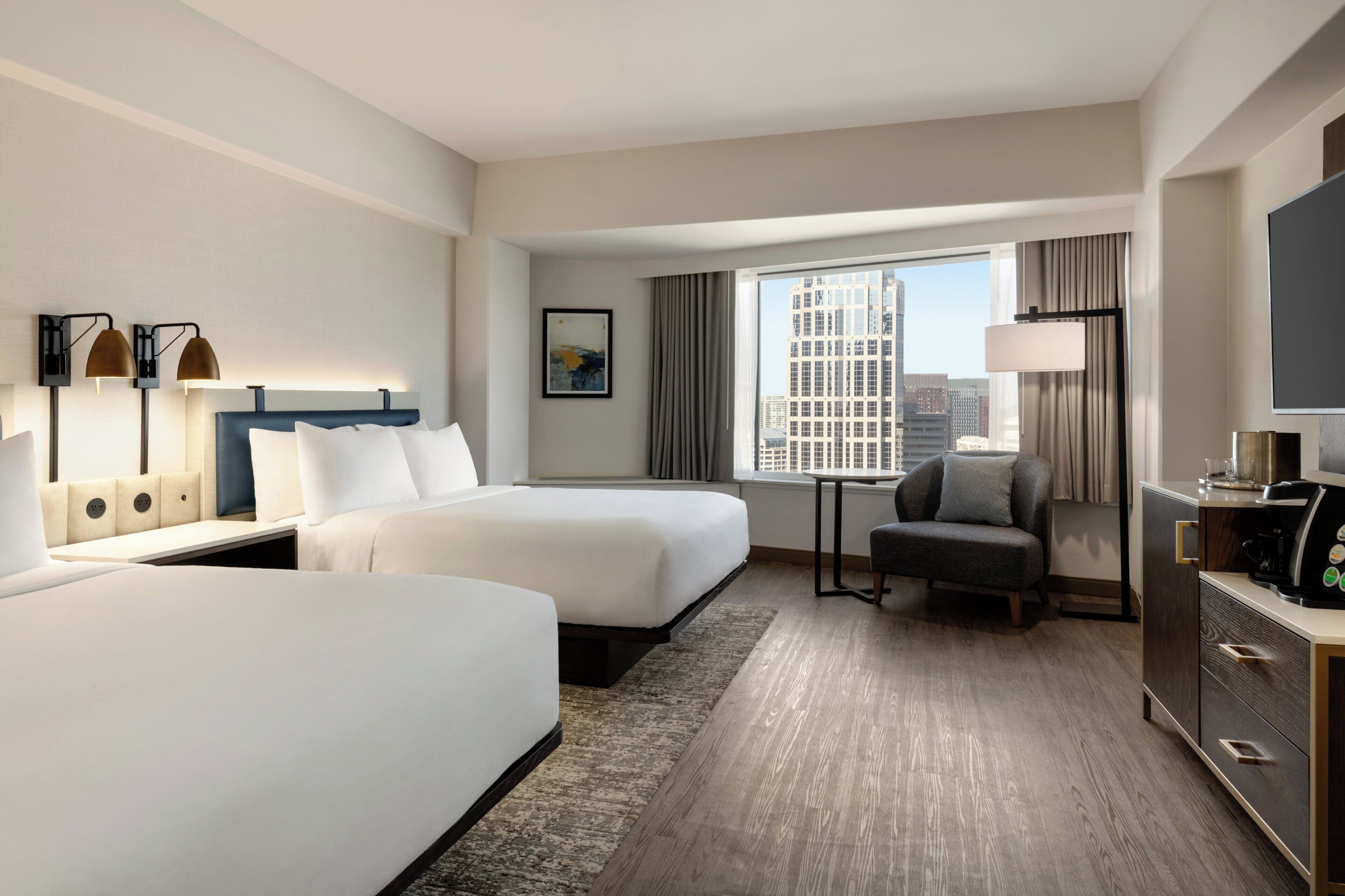 Quarto de hotel com duas camas, uma mesa de trabalho, uma cadeira, televisão e uma janela grande com vista para a cidade.