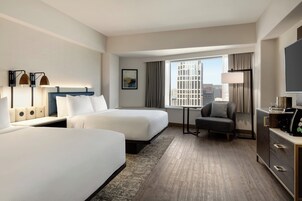 Quarto de hotel com duas camas, uma mesa de trabalho, uma cadeira, televisão e uma janela grande com vista para a cidade.
