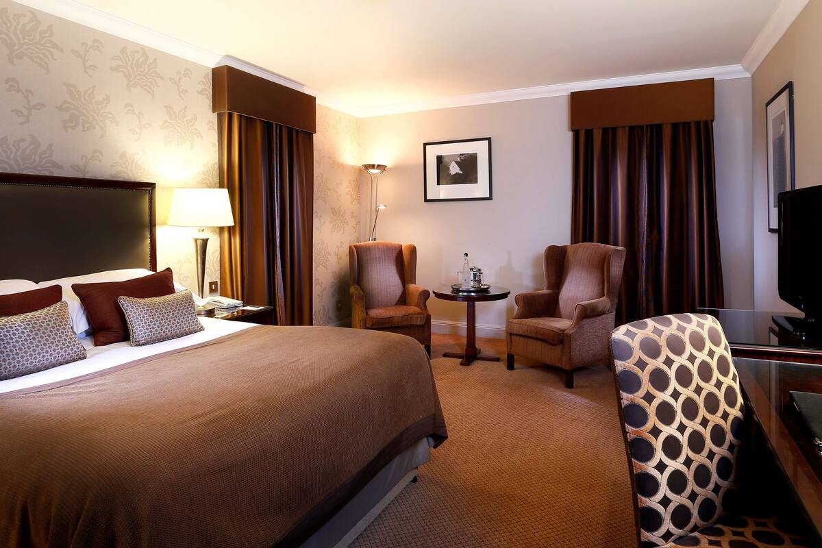 Quarto de hotel com uma cama grande, duas poltronas, uma mesinha, uma luminária e uma televisão.