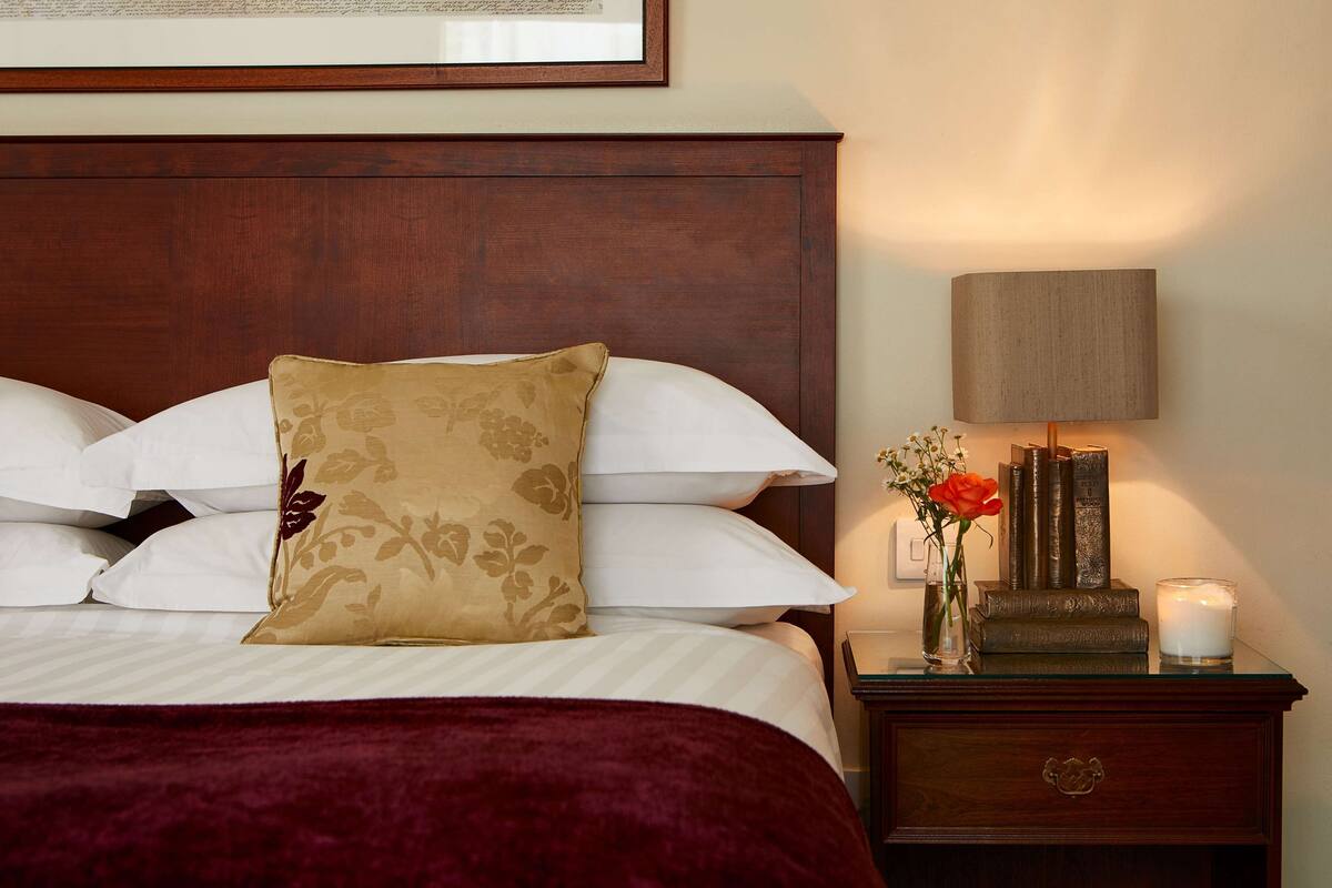 Uma cama bem arrumada com almofadas brancas e bege, um cobertor vermelho e uma cabeceira de madeira. Uma mesa de cabeceira com uma luminária, um vaso com flores e uma vela estão visíveis.