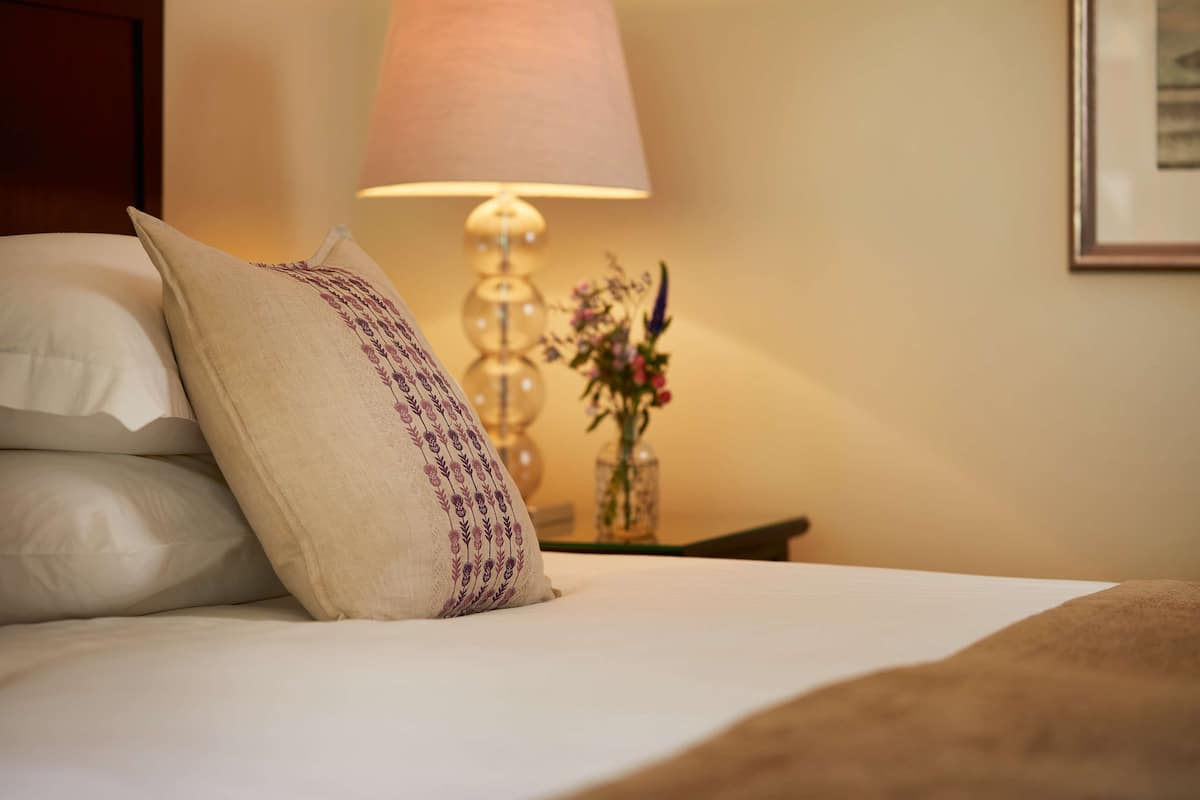Cama bem arrumada com almofadas brancas e bege, abajur decorativo e um vaso com flores sobre uma mesa de cabeceira.