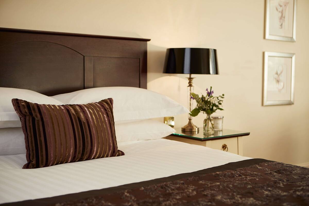 Cama bem arrumada com almofadas listradas, cabeceira escura, mesa de cabeceira com abajur e quadro na parede.