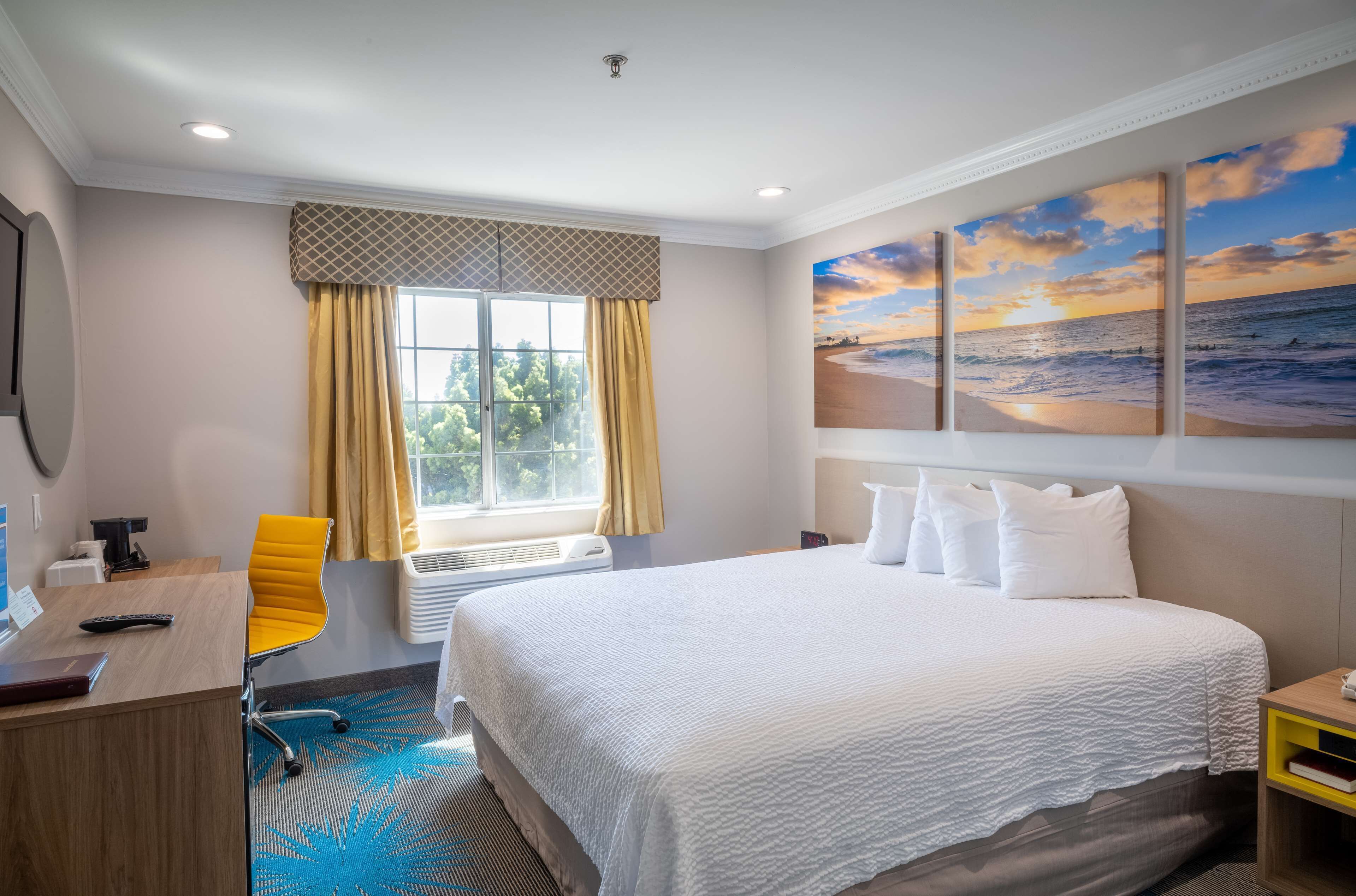 Quarto de hotel com uma cama grande, uma mesa com cadeira, uma janela com cortinas e um quadro de uma cena de praia.