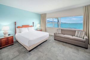 Quarto de hotel com cama, sofá, mesa de cabeceira, luminária e janela com vista para o mar.