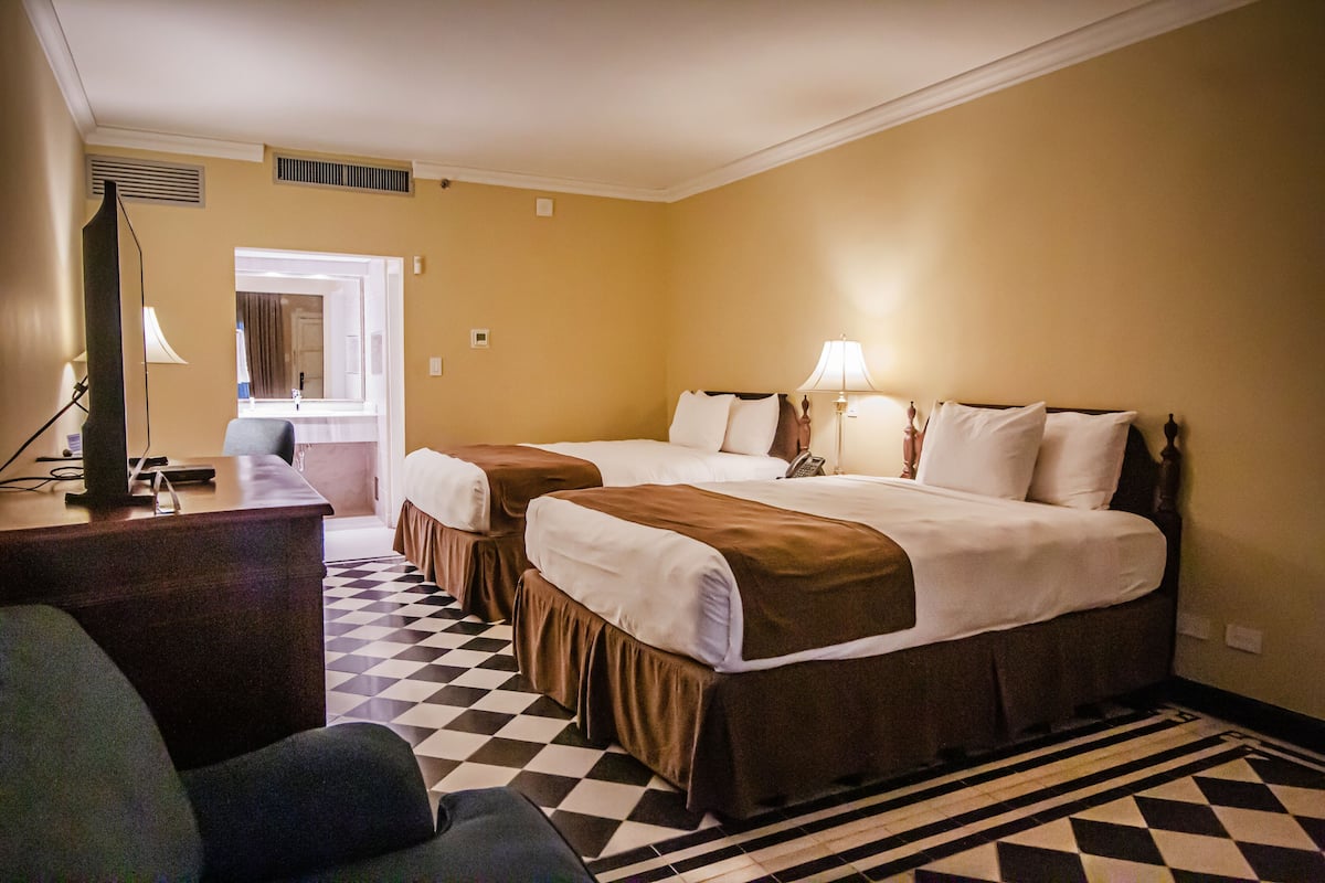 Quarto de hotel com duas camas, chão xadrez, mesa com luminária e janela com cortinas.
